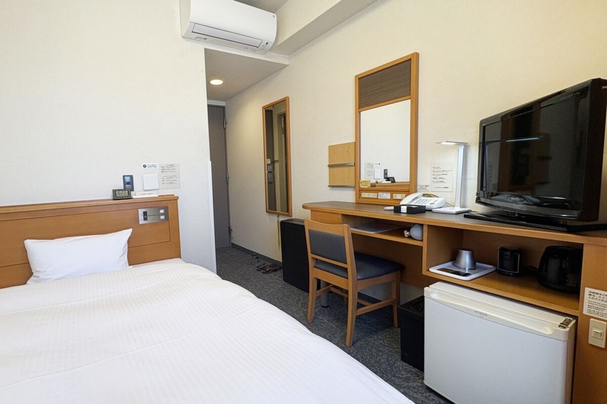 Quarto de hotel com cama, escrivaninha com cadeira, televisão, mini-geladeira e micro-ondas.