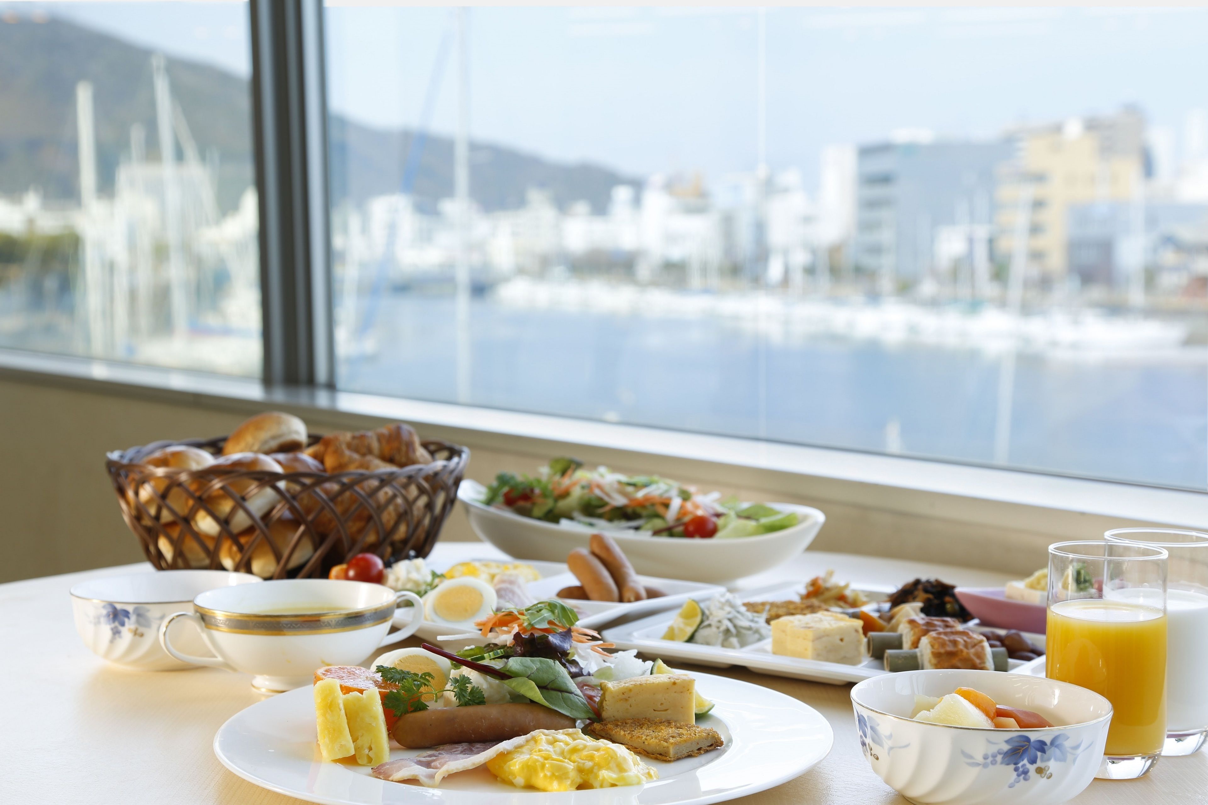 Uma mesa de café da manhã com diversos pratos, uma salada e vista para uma marina com barcos e edifícios.
