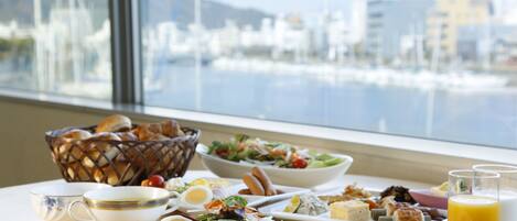 Uma mesa de café da manhã com diversos pratos, uma salada e vista para uma marina com barcos e edifícios.