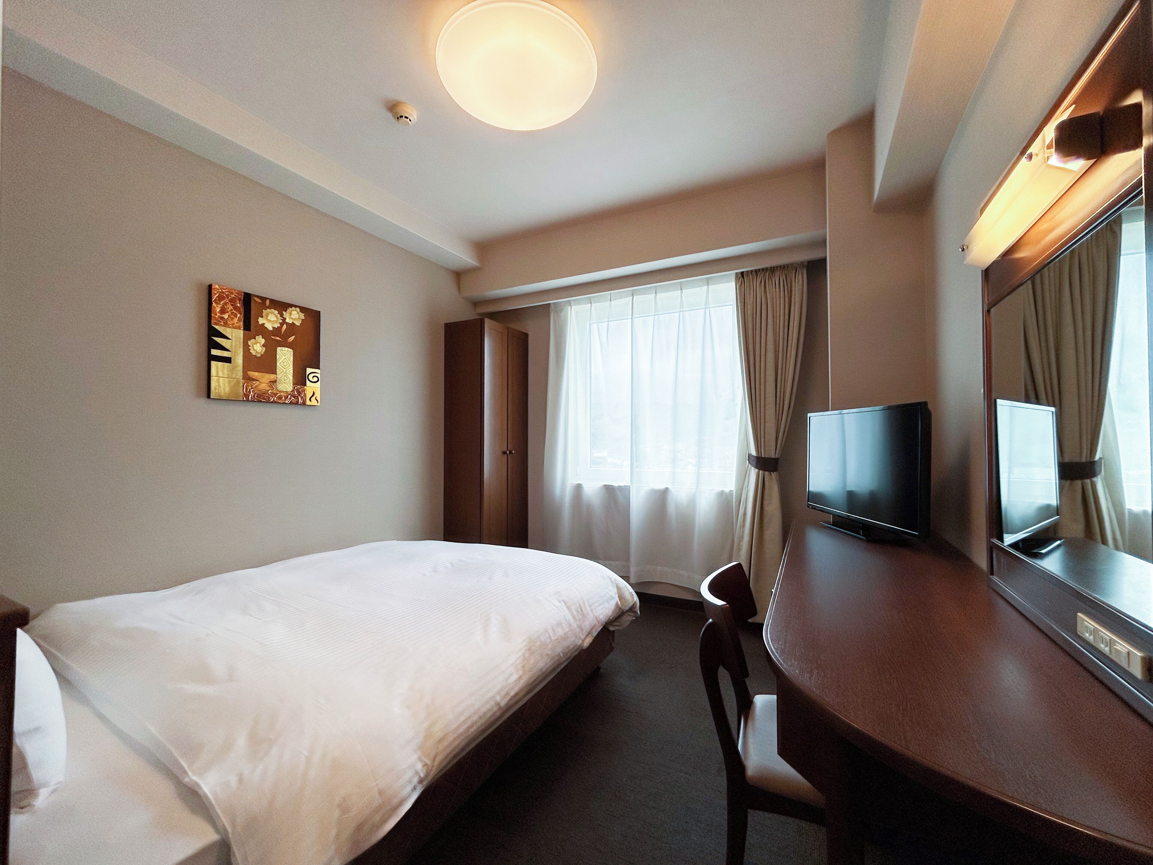 Quarto de hotel com cama, escrivaninha com televisão, cadeira, guarda-roupa e janela com cortinas.