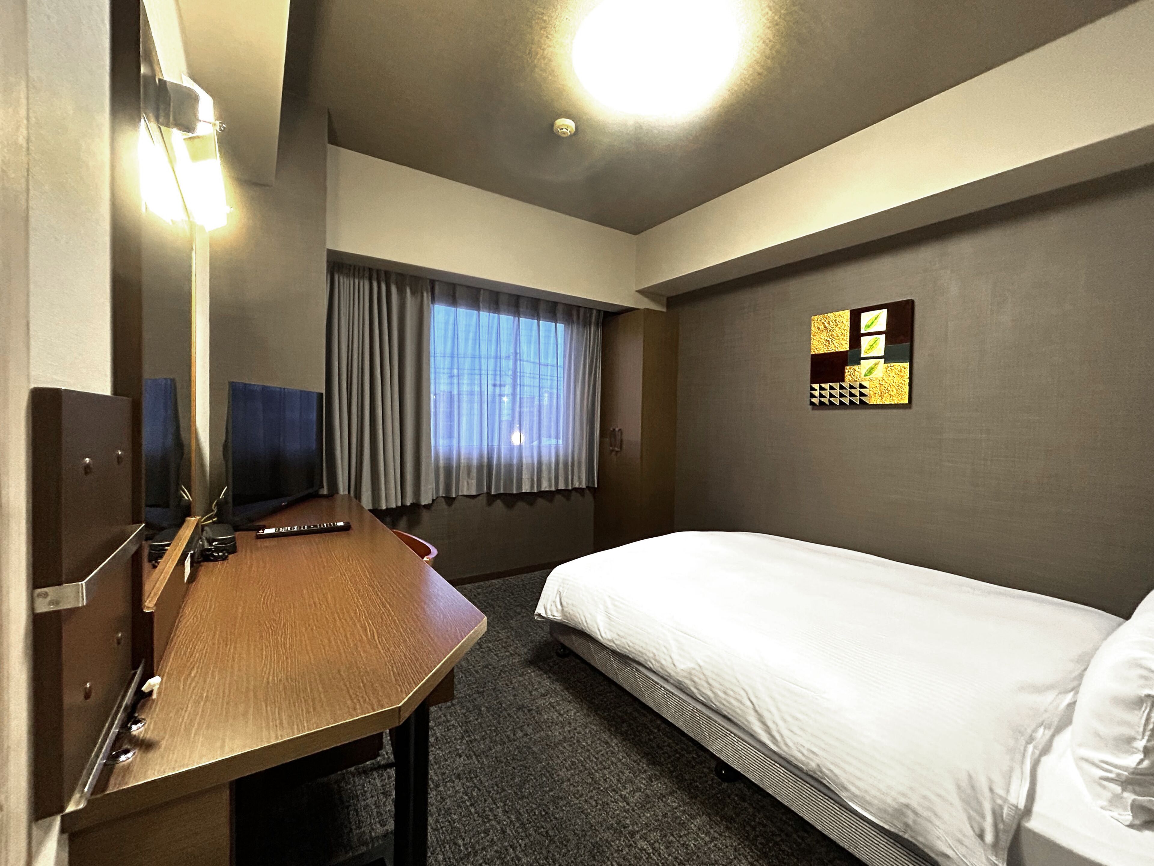 Quarto de hotel com cama, escrivaninha com cadeira, televisão e janela com cortinas.
