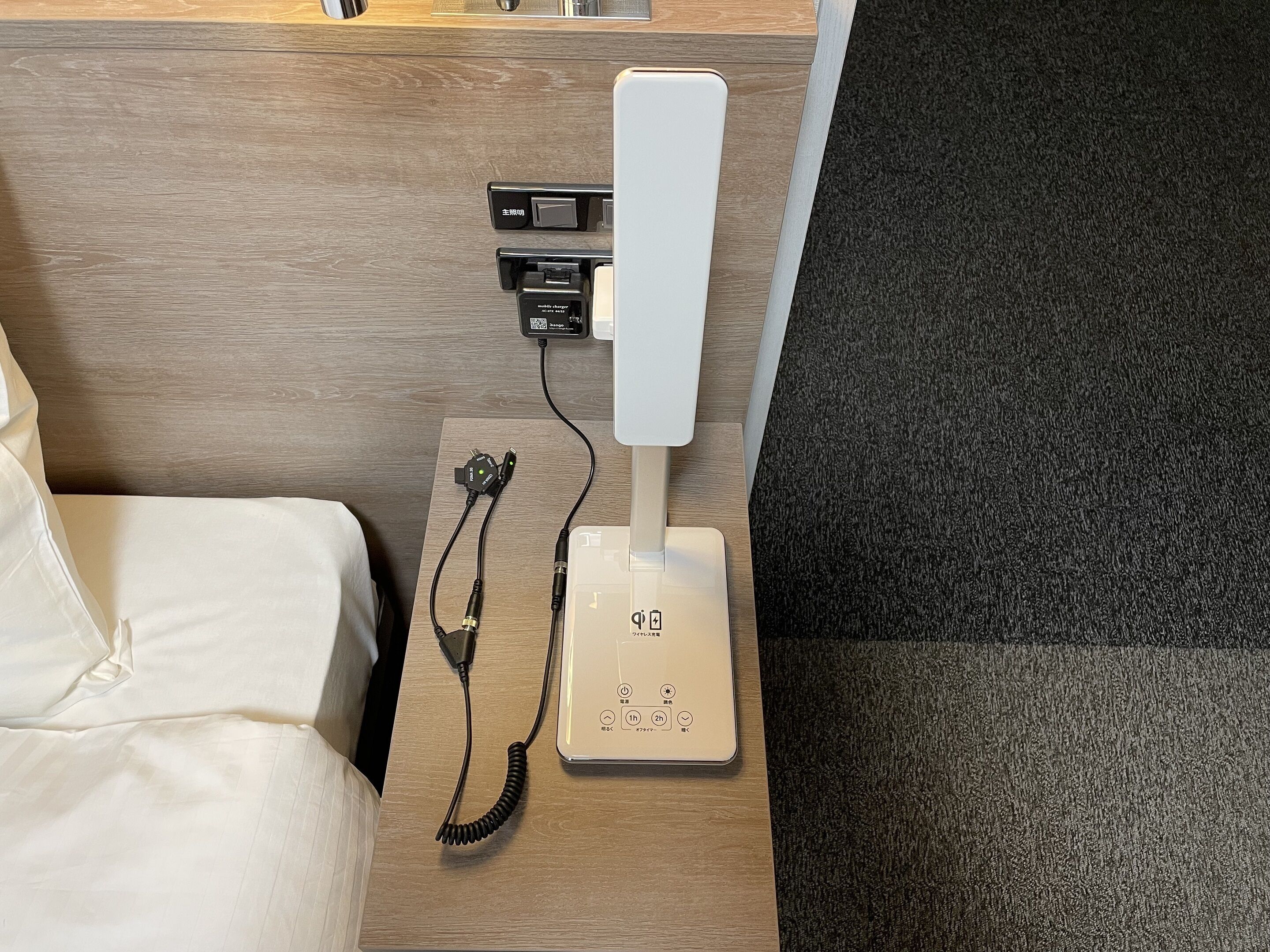 Quarto de hotel com mesa de cabeceira, abajur branco, carregador de celular e tomada.