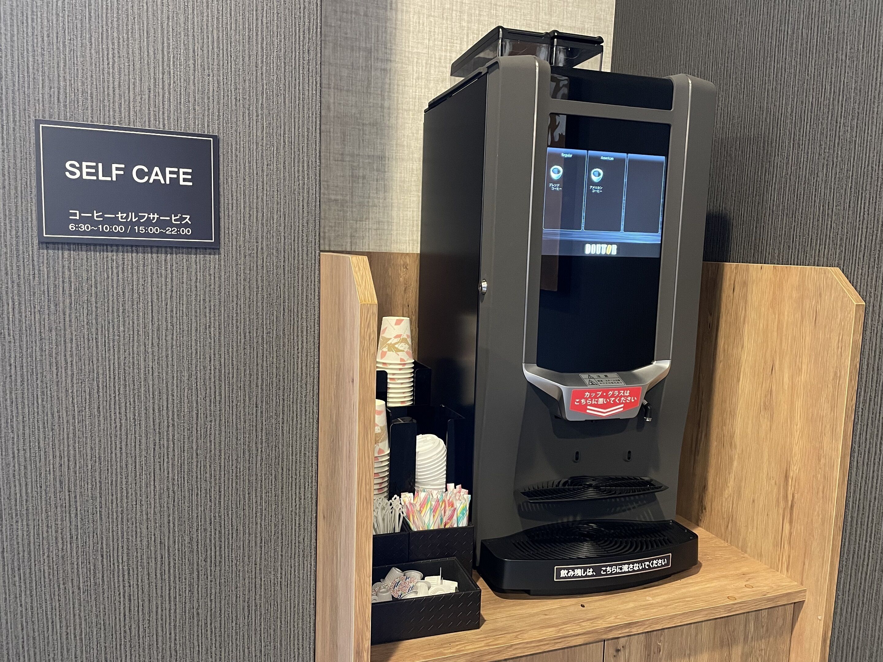 Uma máquina de café automática com visor digital, uma pilha de copos de papel e uma pequena lixeira.