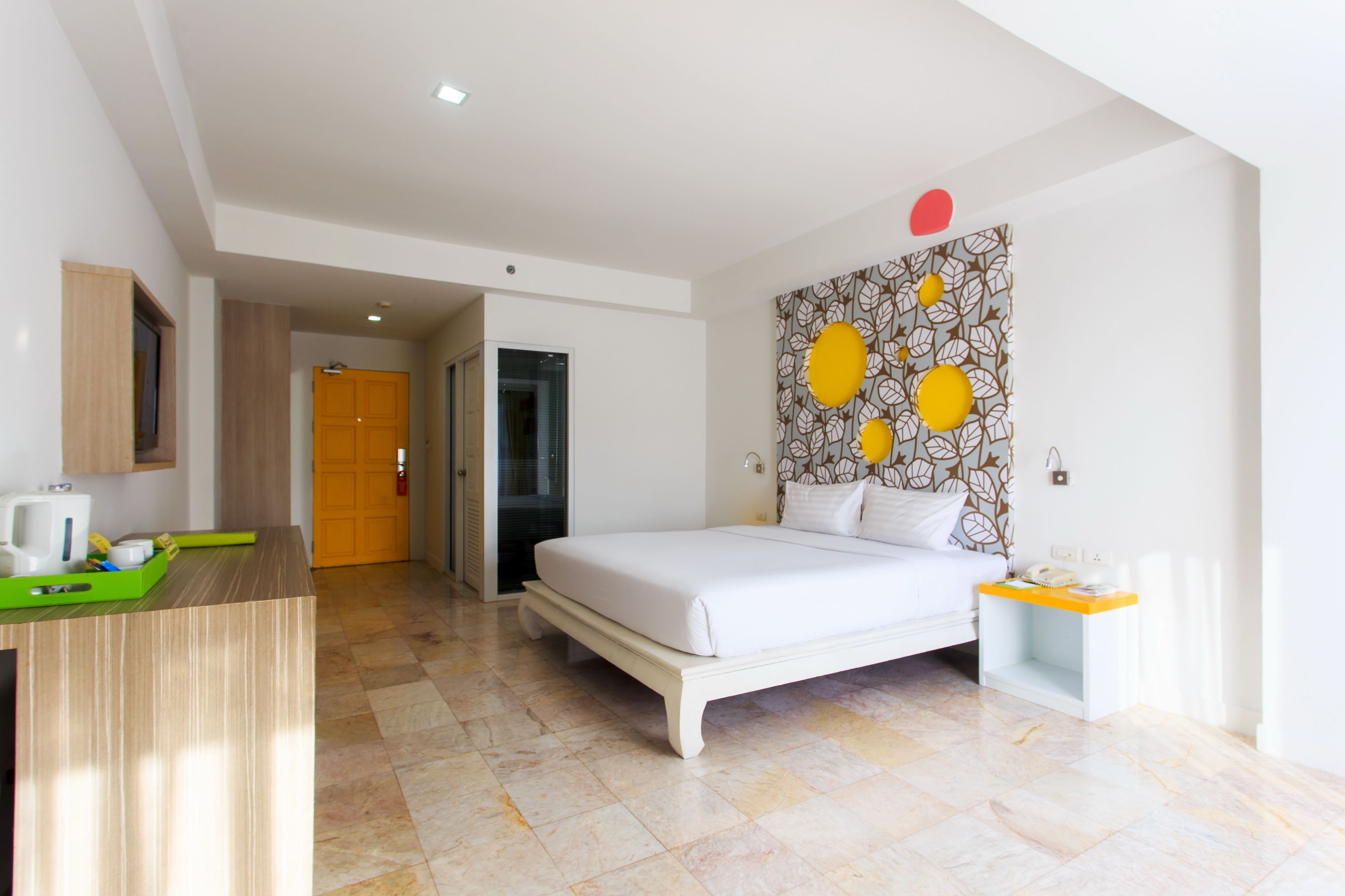 Quarto de hotel moderno com uma cama grande, uma escrivaninha com um tabuleiro verde, uma televisão na parede e uma porta que dá para outro quarto.