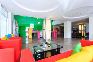 Um lobby moderno com paredes verde-claras, assentos vermelhos e amarelos, e uma mesa de centro de vidro com relógio e planta.