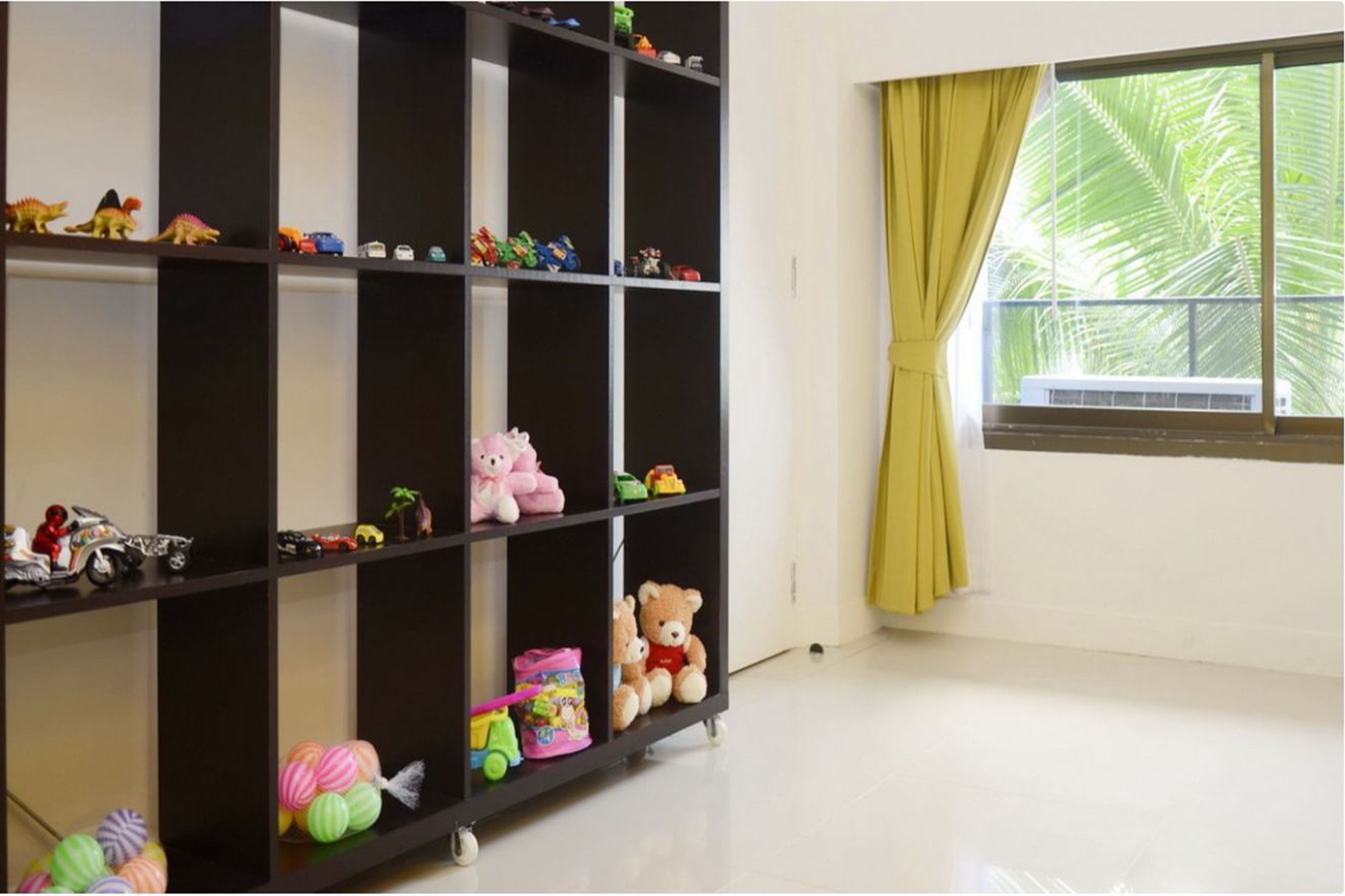 Quarto com uma estante preta cheia de brinquedos e uma janela com cortinas amarelas.