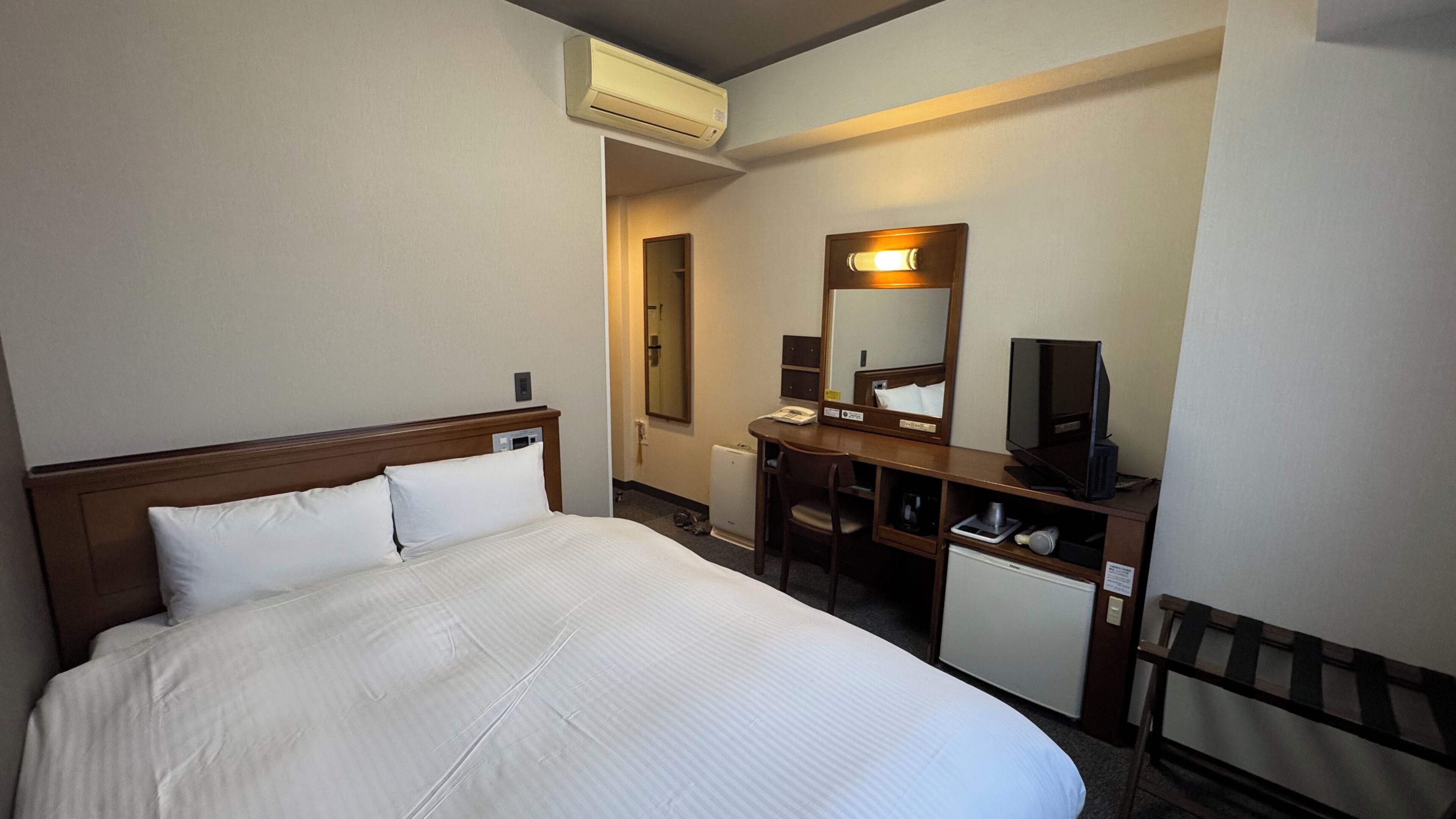 Quarto de hotel com cama, escrivaninha, cadeira, televisão e ar condicionado.
