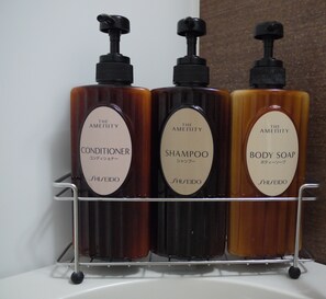Três frascos dos produtos de higiene pessoal da marca The Amenity sobre uma prateleira de metal.