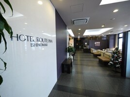 Uma área de recepção de hotel com uma placa indicando "HOTEL ROUTE INN ISHINOMAKI". Há áreas de estar com um pinheiro de Natal e um balcão de recepção.