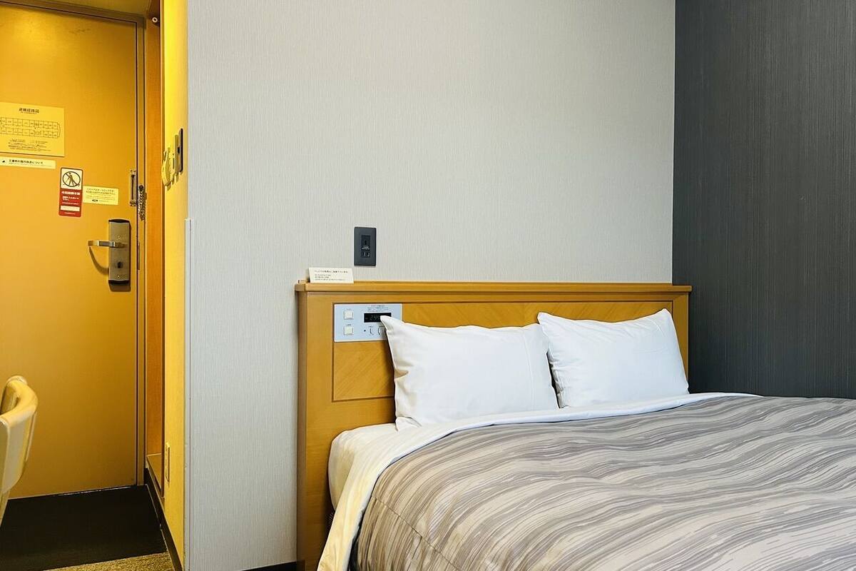 Quarto de hotel com uma cama, cabeceira de madeira, painel de controle e uma porta com sinalização.