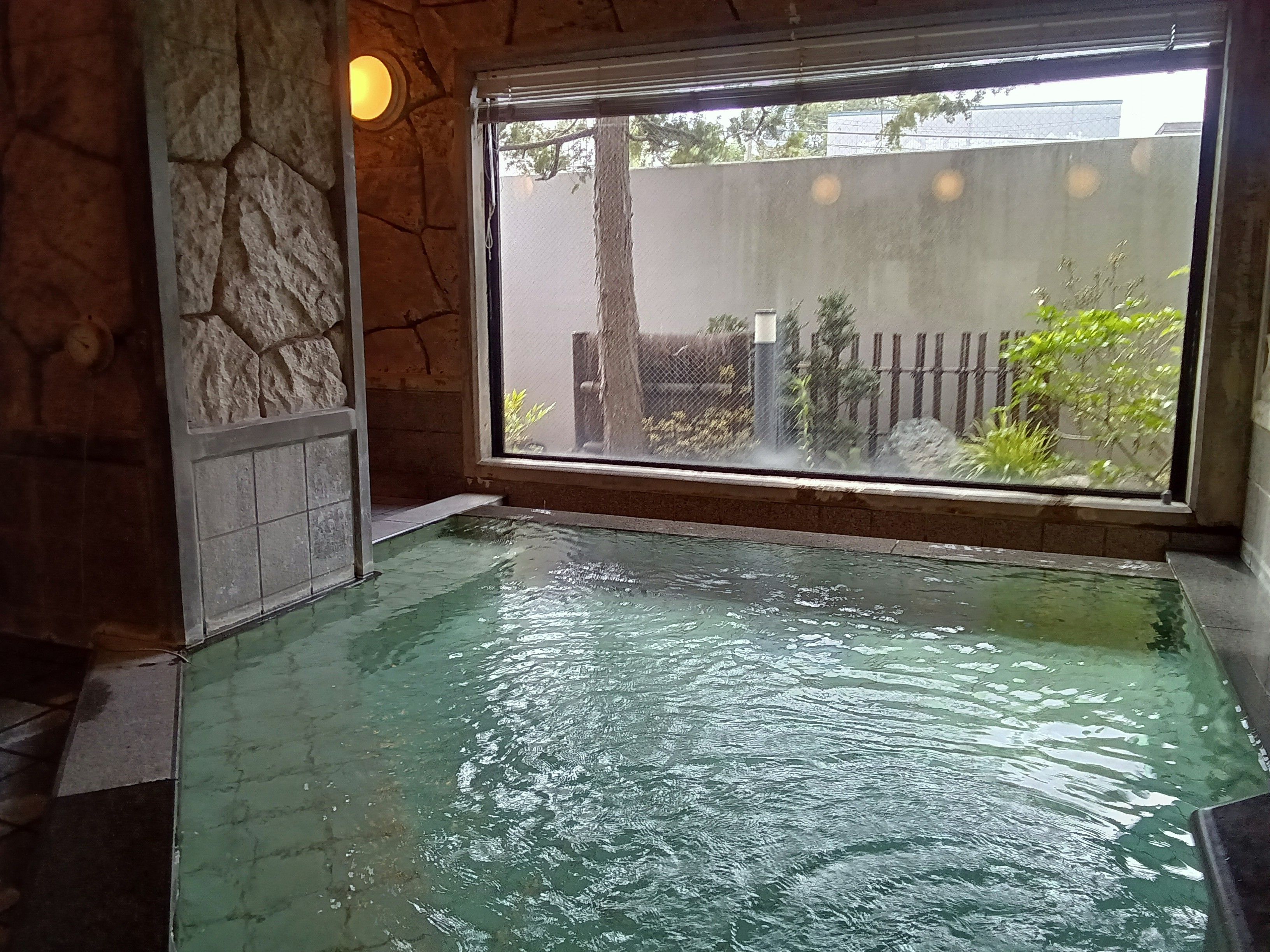 Área de spa com parede de pedra, janela com vista para cerca e vegetação, e uma pequena piscina.
