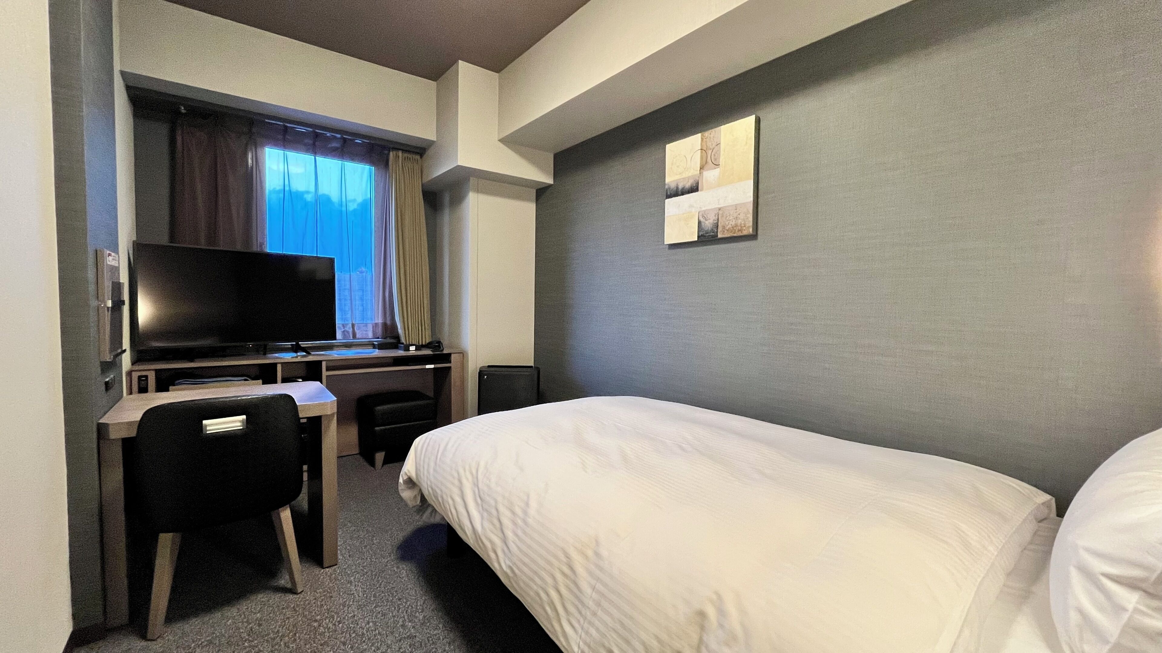 Quarto de hotel com cama, escrivaninha com computador, televisão e janela com cortinas.