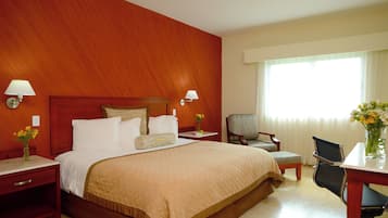 Quarto de hotel com uma cama grande, duas mesas de cabeceira com luminárias, uma escrivaninha com cadeira e uma janela com cortinas.