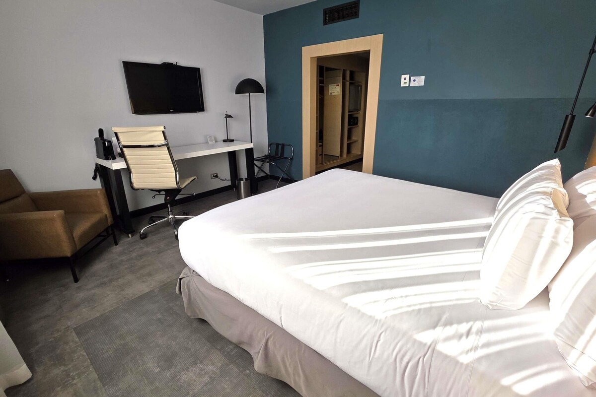 Quarto de hotel com cama, escrivaninha, cadeira, televisão e porta.