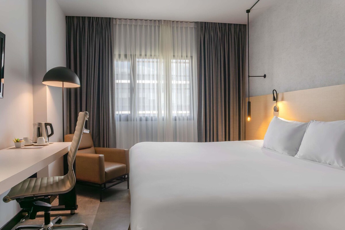 Quarto de hotel com uma cama, uma mesa de escritório com cadeira, luminária e janela com cortinas.
