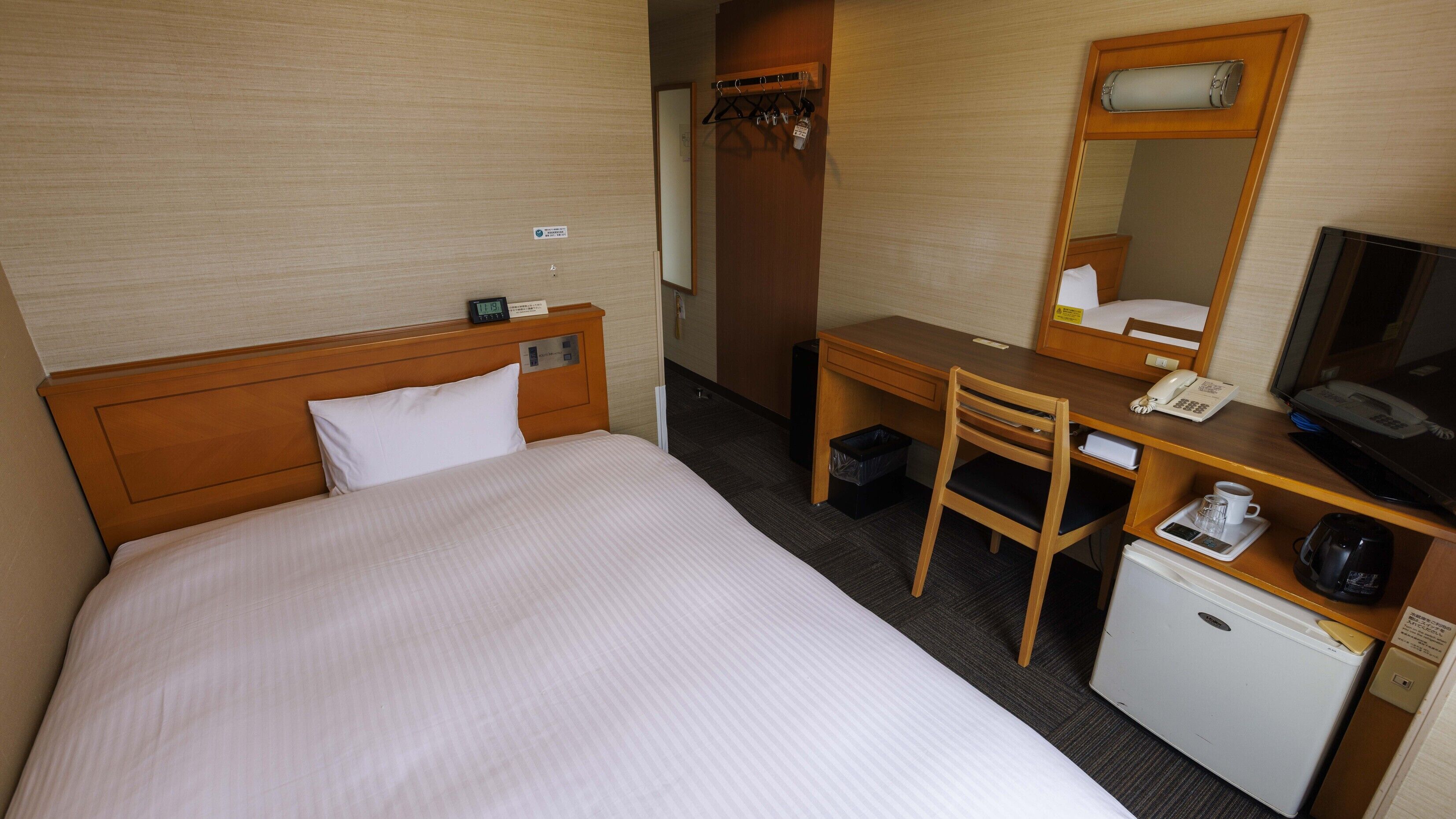 Quarto de hotel com cama, escrivaninha, cadeira, pequeno frigorífico, espelho, televisão e relógio.