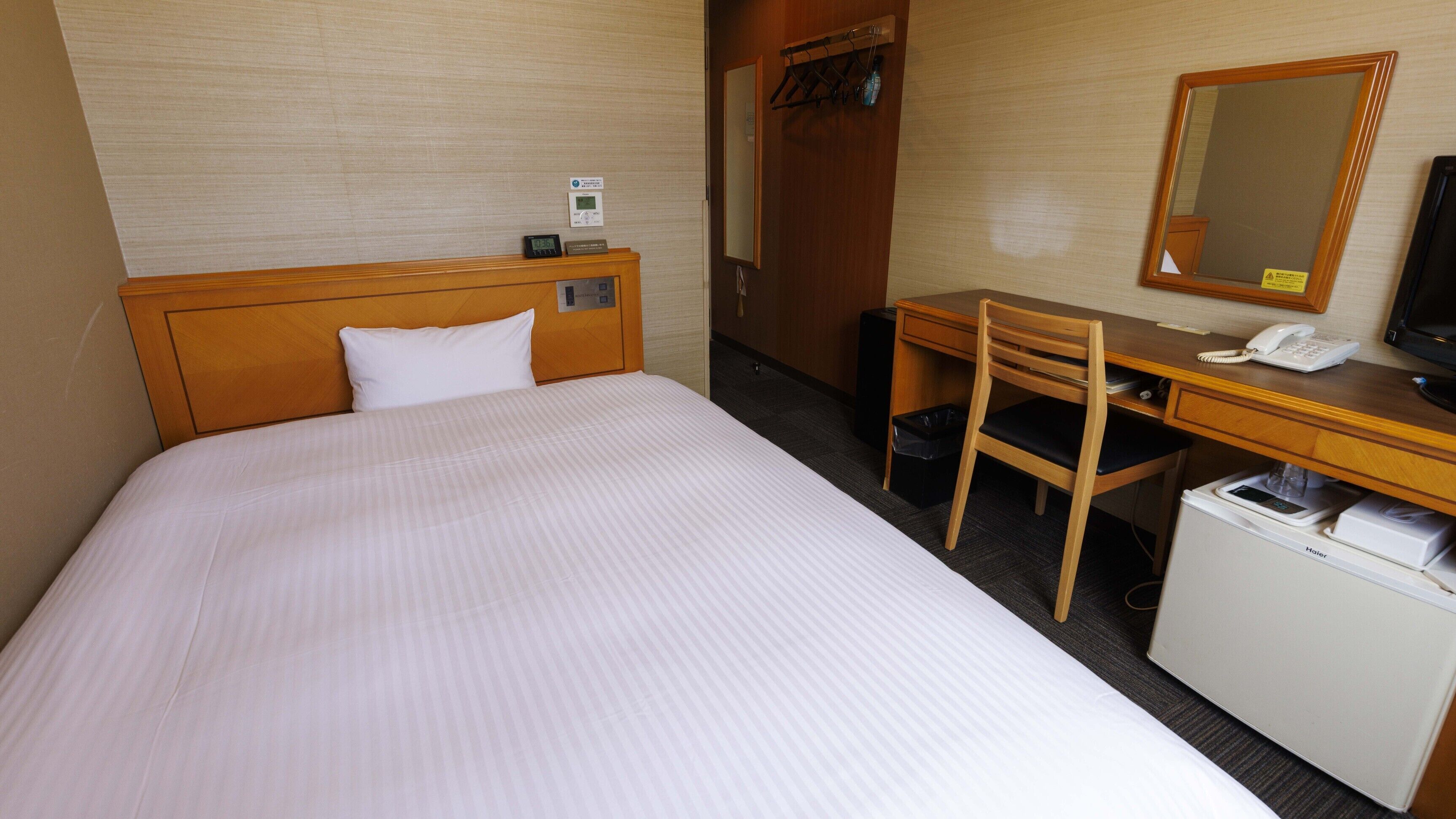 Quarto de hotel com cama, escrivaninha, cadeira, telefone, espelho e um pequeno frigorífico.