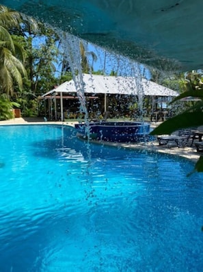 Piscina azul cristalina com hidromassagem, cercada por palmeiras e uma área coberta com mesas e guarda-sóis.