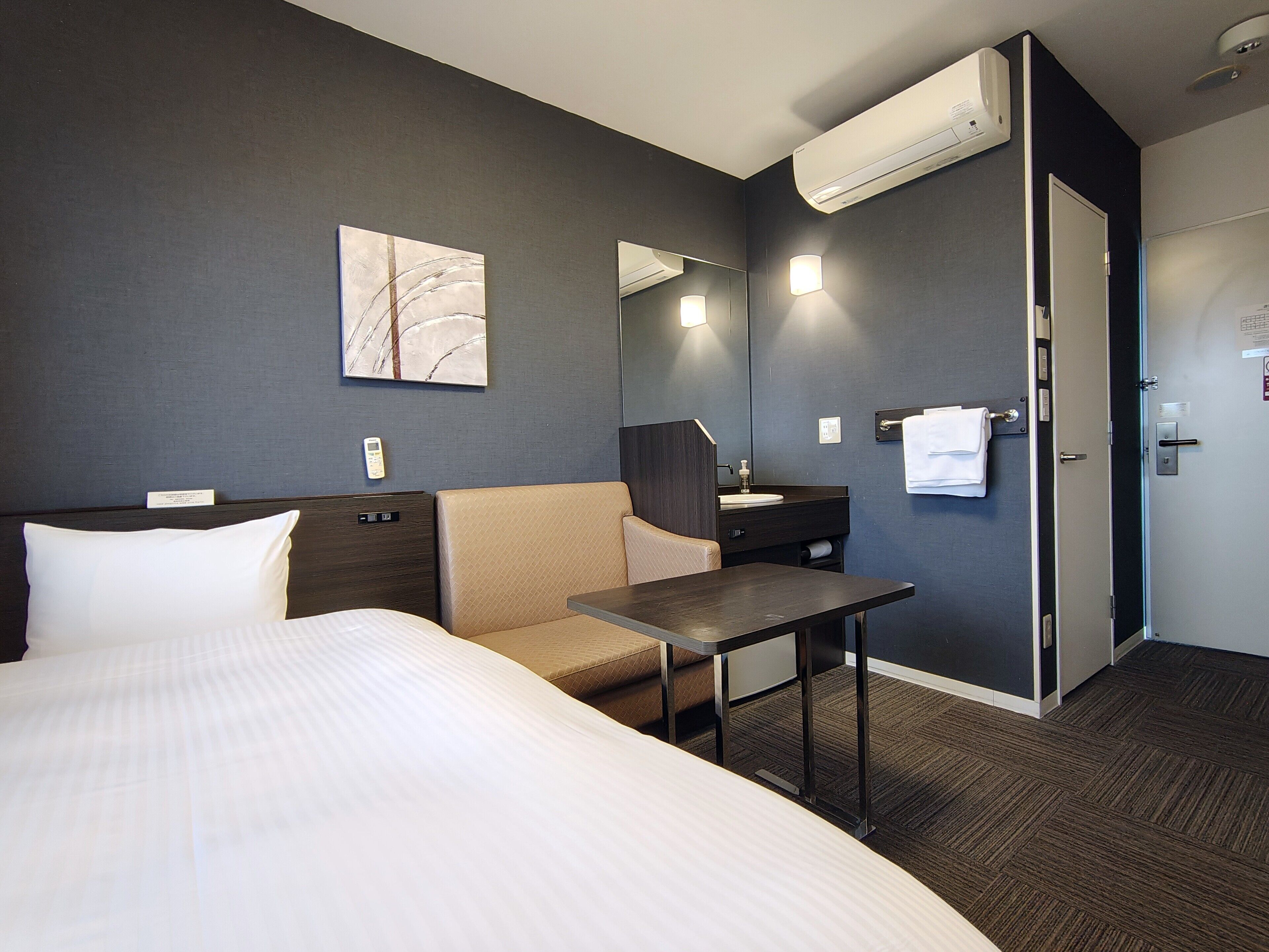 Quarto de hotel com cama, mesa pequena, sofá, escrivaninha, espelho, pia e aparelho de ar condicionado.