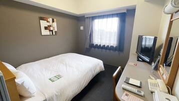 Quarto de hotel com uma cama de solteiro, escrivaninha com telefone, televisão e janela com cortinas.