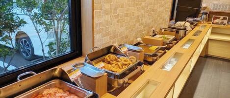 Uma área de buffet com diversas opções de comida, incluindo bacon, anéis de cebola e outros pratos, dispostos em um balcão com parede de tijolos ao fundo.