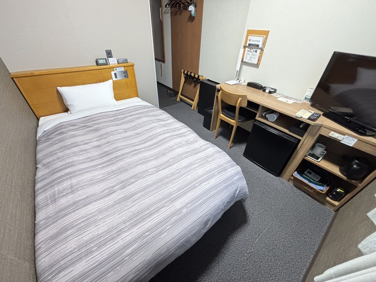 Quarto de hotel com cama, escrivaninha com computador, cadeira, televisão e pequeno frigorífico.