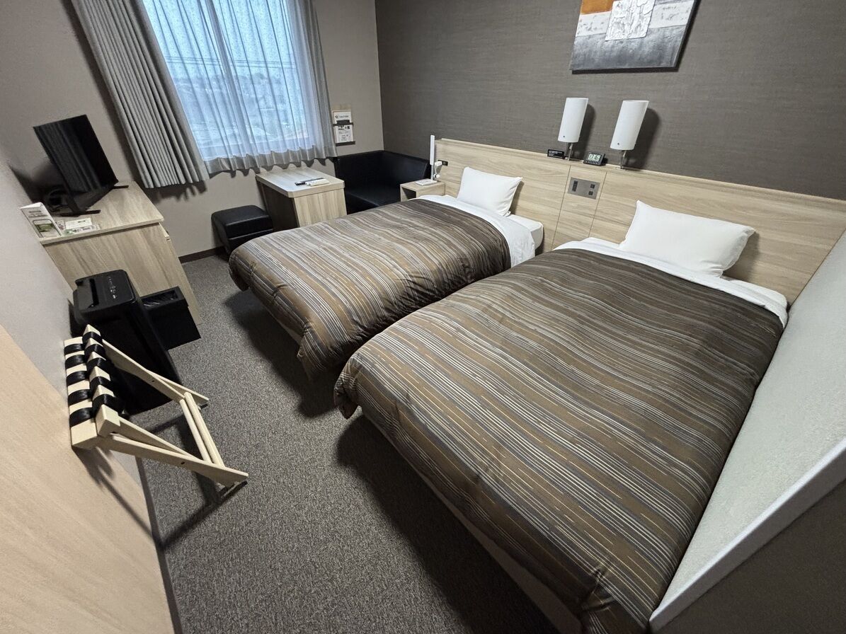 Quarto de hotel com duas camas, uma escrivaninha, uma cadeira, televisão e janela com cortinas.