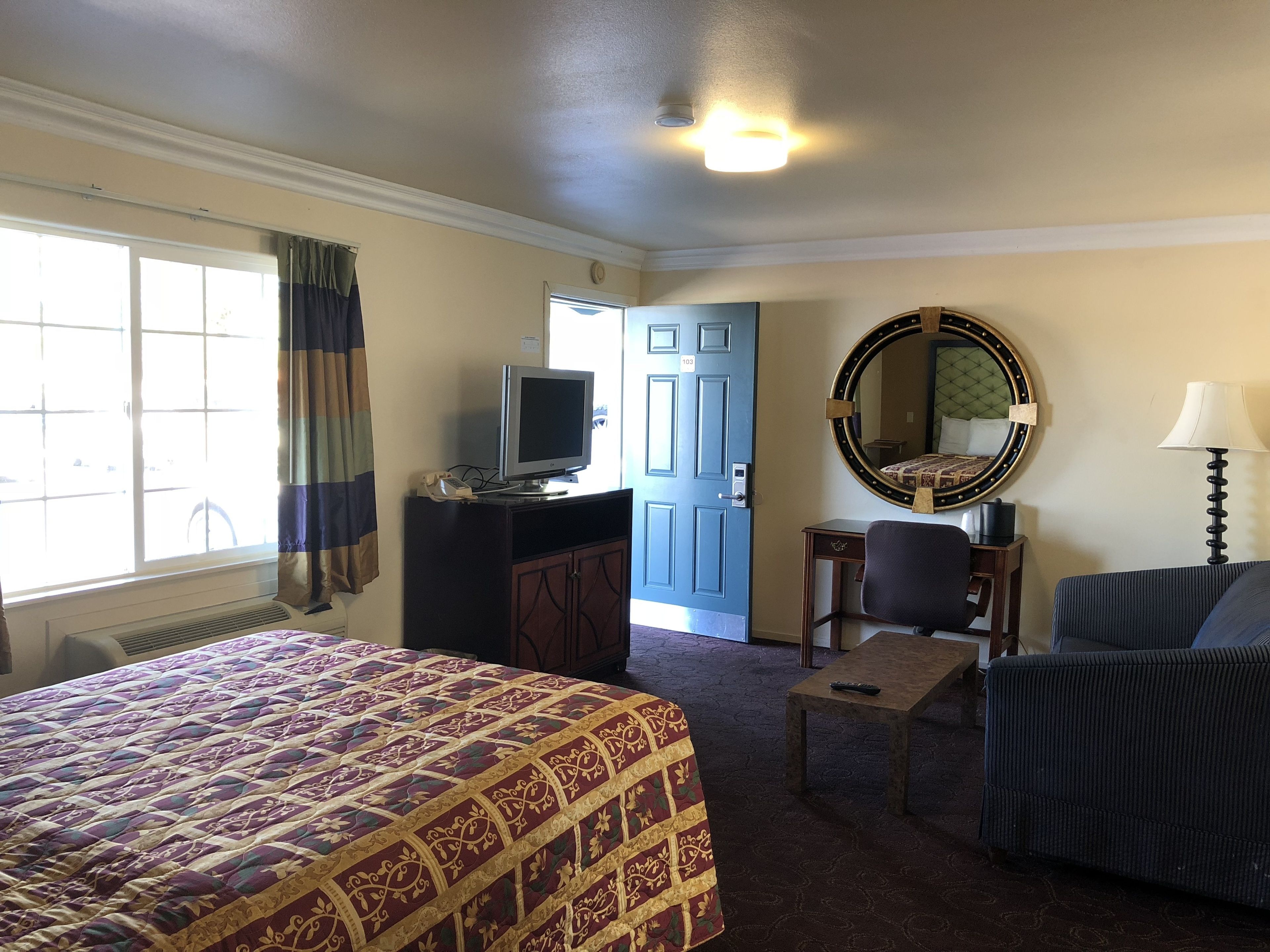 Quarto de hotel com uma cama grande, uma escrivaninha com televisão, uma cadeira, um espelho redondo e um abajur.