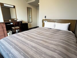 Quarto de hotel com uma cama grande, uma escrivaninha com cadeira, um espelho e uma mesinha de cabeceira.