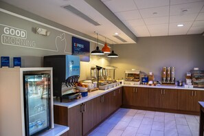 Espaço moderno para café da manhã com geladeira, máquinas de café e diversas opções de alimentos.