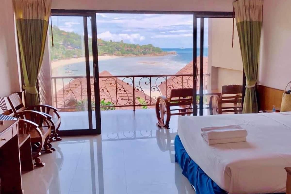 Quarto de hotel com cama, escrivaninha e cadeira. Tem uma varanda com vista para a praia e o mar.