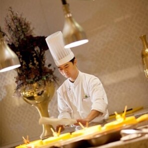 Um chef com uniforme branco e chapéu alto, trabalhando em uma bancada de cozinha com uma panela grande e um vaso decorativo ao fundo.