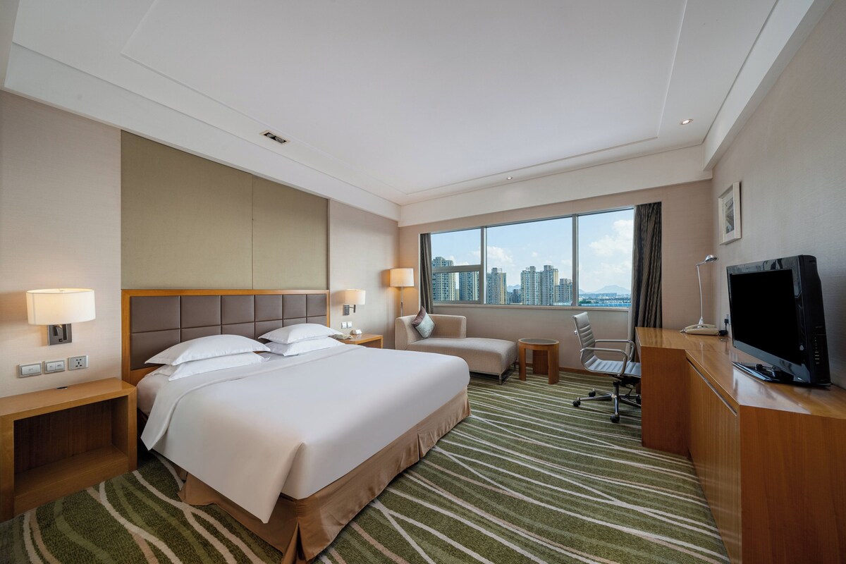 Quarto de hotel com uma cama grande, uma escrivaninha e televisão, e vista para a paisagem urbana.