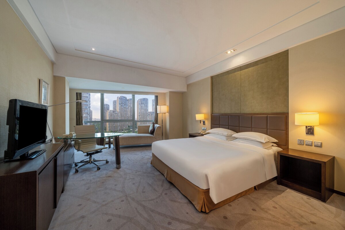 Quarto de hotel com cama, mesa de cabeceira, luminária, escrivaninha com cadeira, televisão e vista da cidade através de amplas janelas.