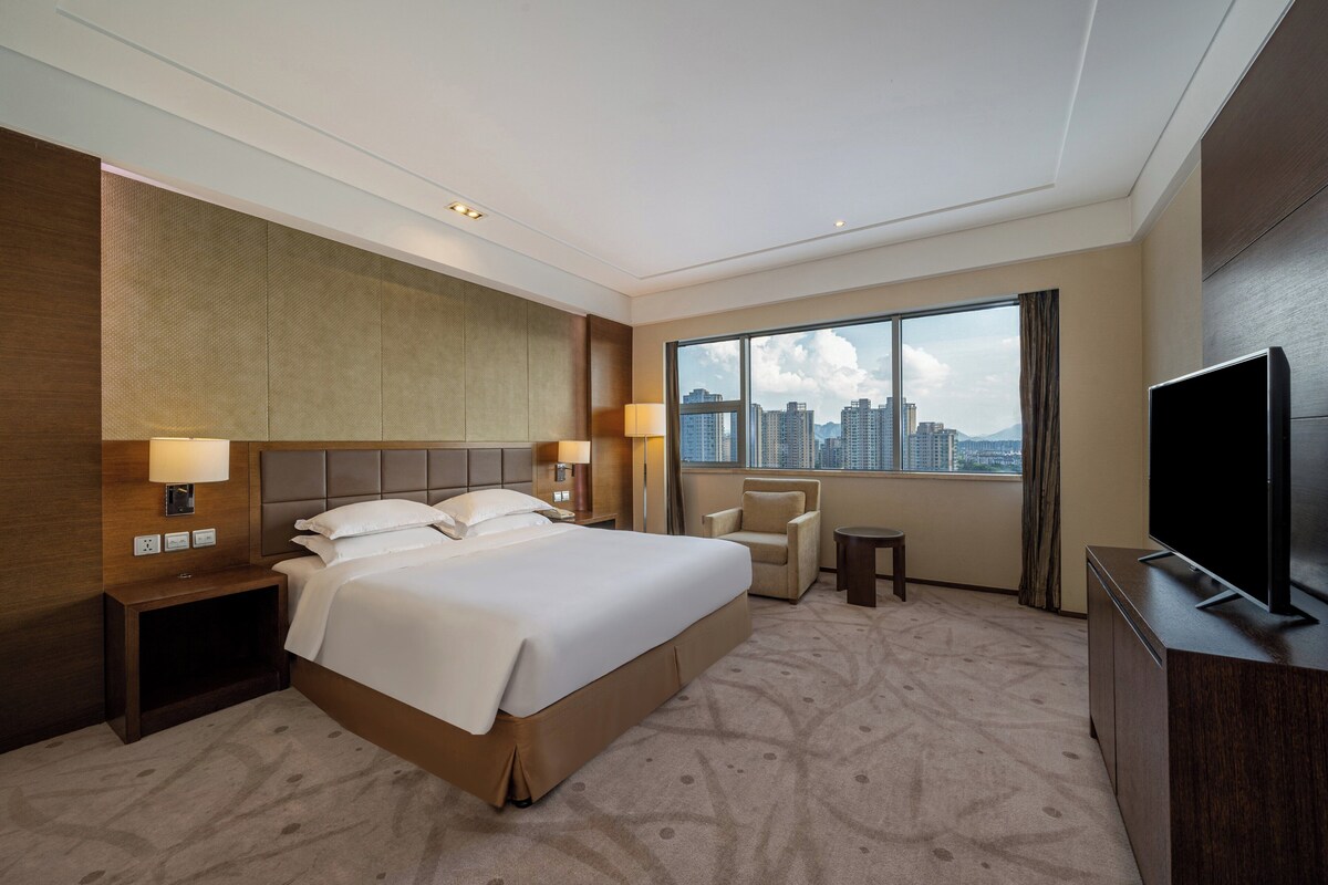 Quarto de hotel com uma cama grande, televisão, poltrona e vista para a paisagem urbana.