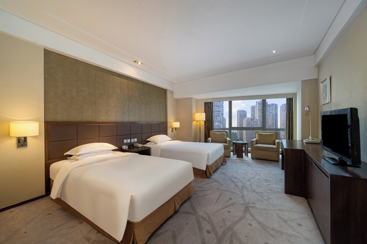 Quarto de hotel com duas camas, televisão, escrivaninha e vista para a cidade.