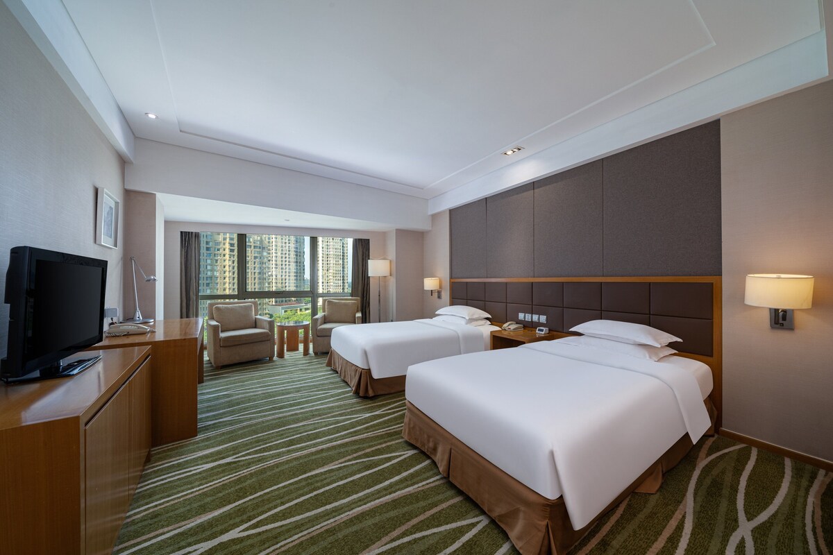Quarto de hotel com duas camas, uma escrivaninha com televisão, uma cadeira e vista para a cidade.