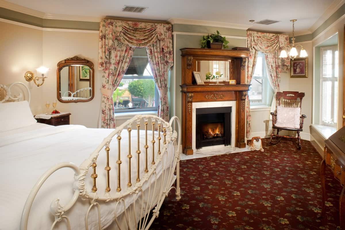 Quarto com cama grande, lareira, janela com cortinas e uma cadeira.