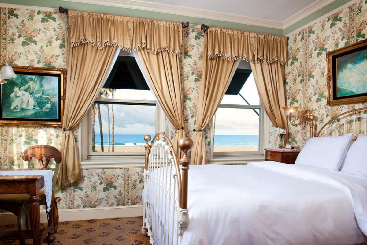 Quarto com uma cama grande, mesa de cabeceira de madeira, escrivaninha e uma cadeira. Há duas janelas com cortinas e um quadro na parede.