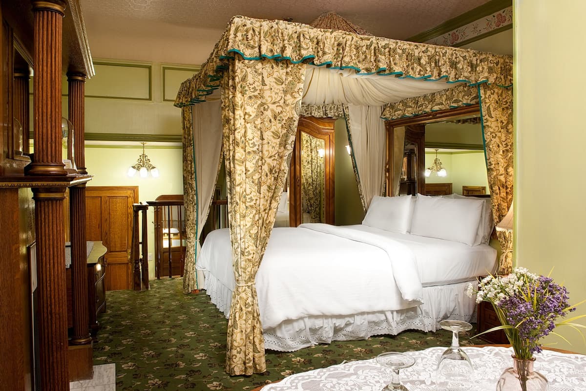 Uma cama com dossel e cortinas florais, um criado-mudo de madeira e um vaso de flores.