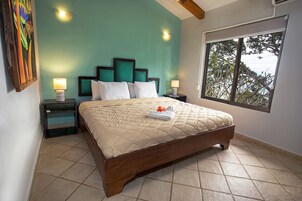 Uma cama bem arrumada com colcha, duas mesas de cabeceira com abajures, uma janela com vista e um quadro na parede.