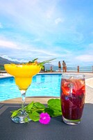 Ambiente à beira da piscina com margarita e um copo de bebida gelada, piscina e vista para o oceano e montanhas ao fundo.