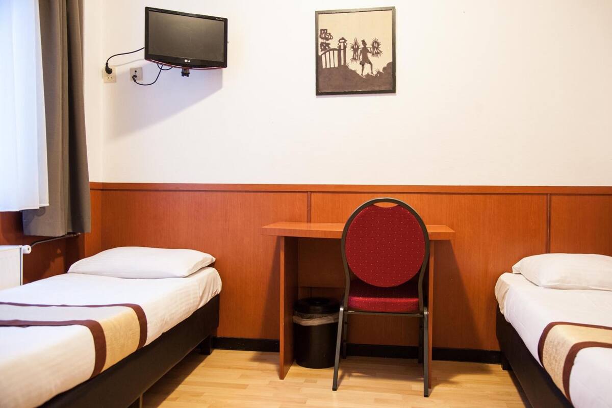 Quarto de hotel com duas camas de solteiro, uma escrivaninha com cadeira, televisão fixada na parede e um quadro sobre a escrivaninha.