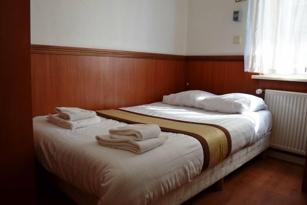 Cama bem arrumada com lençóis brancos e detalhes em marrom, uma cabeceira de madeira e uma janela com cortinas.