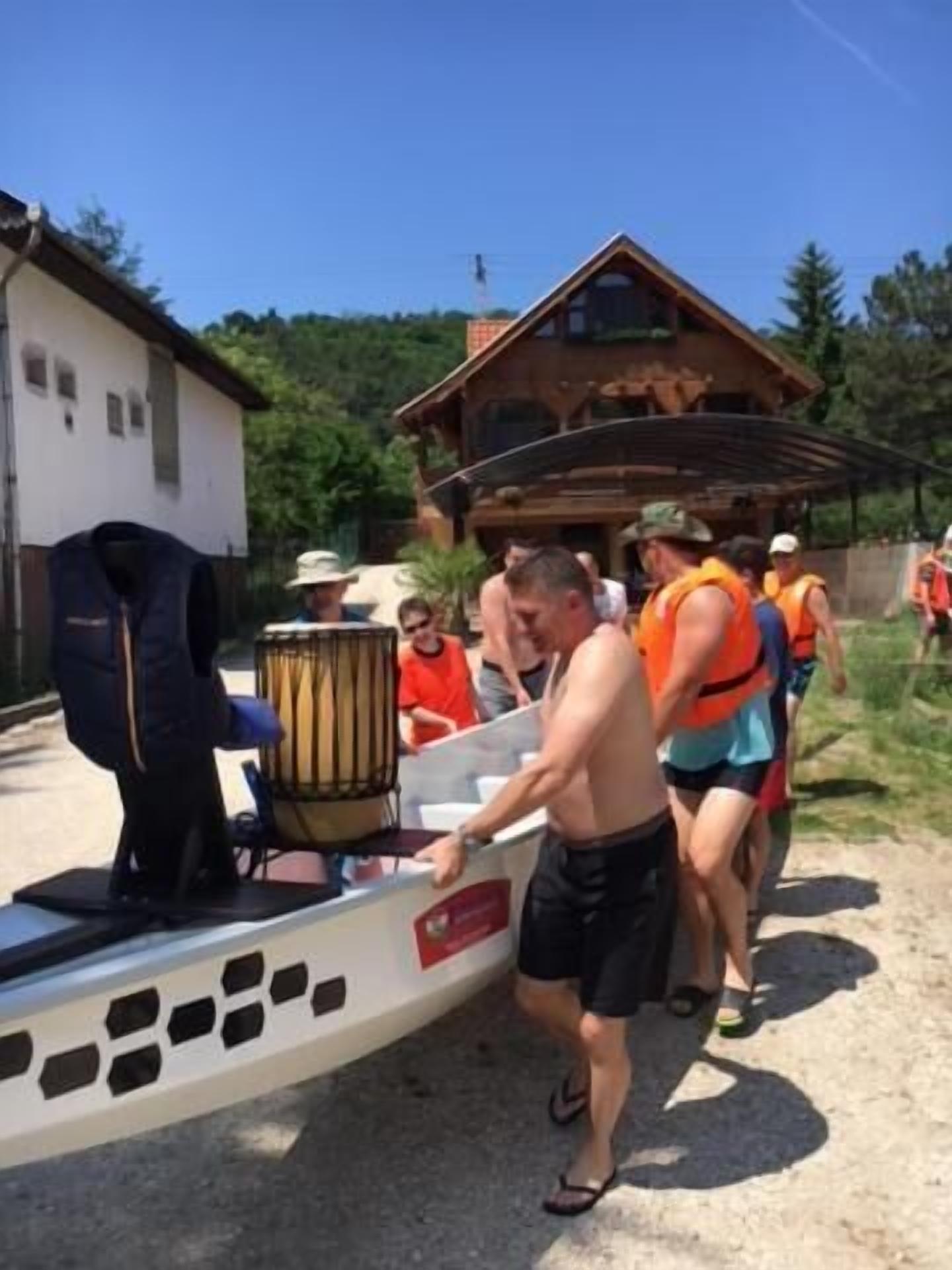 Um grupo de pessoas se preparando para embarcar em um barco, com uma casa e árvores ao fundo.