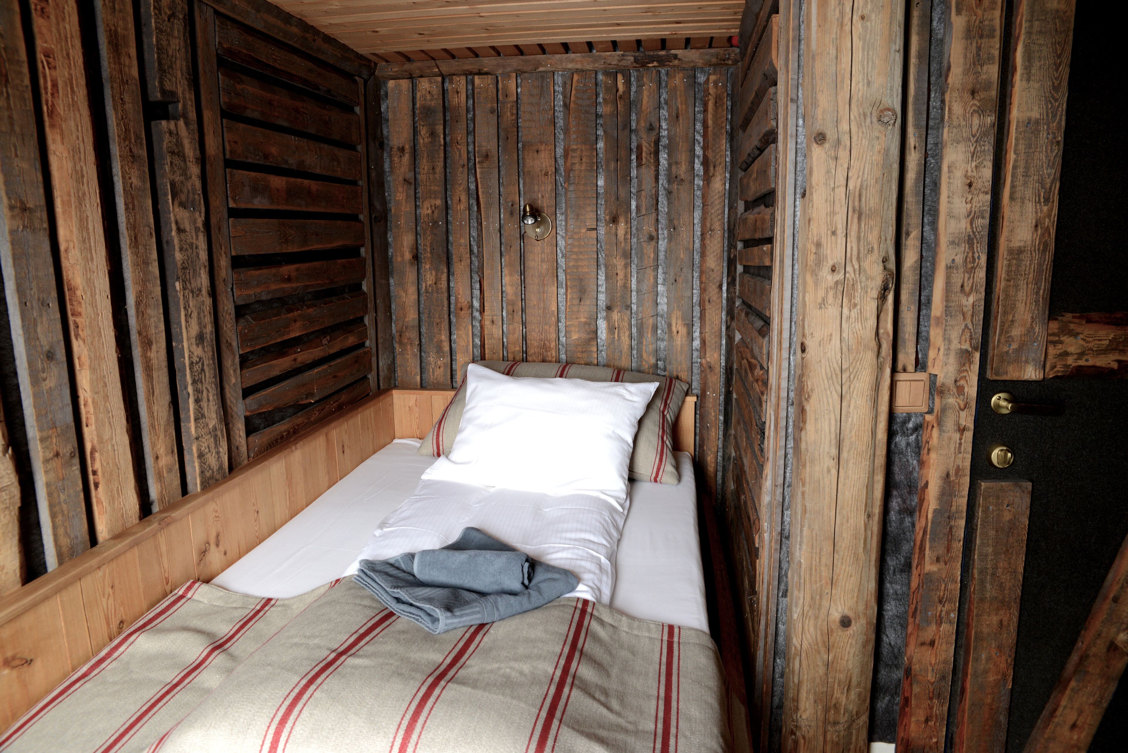 Uma cama pequena com estrutura de madeira, roupa de cama branca e manta listrada.