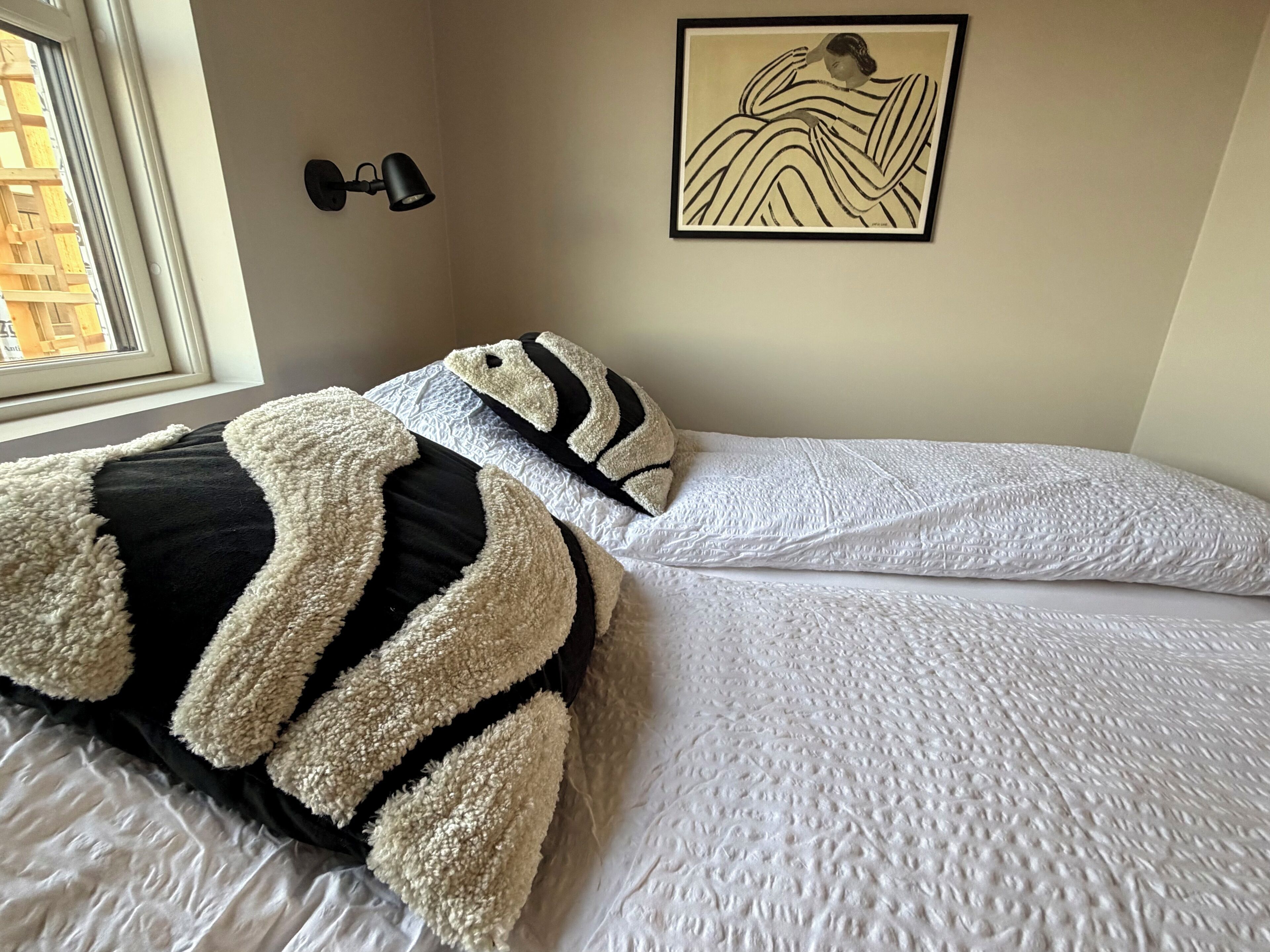 Cama bem arrumada com um cobertor listrado e travesseiro branco, uma obra de arte emoldurada na parede, e uma janela com vista para um prédio de tijolos.
