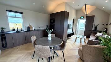Uma cozinha moderna com armários escuros, uma área de jantar com mesa e cadeiras redondas, e uma sala de estar com sofá e planta.