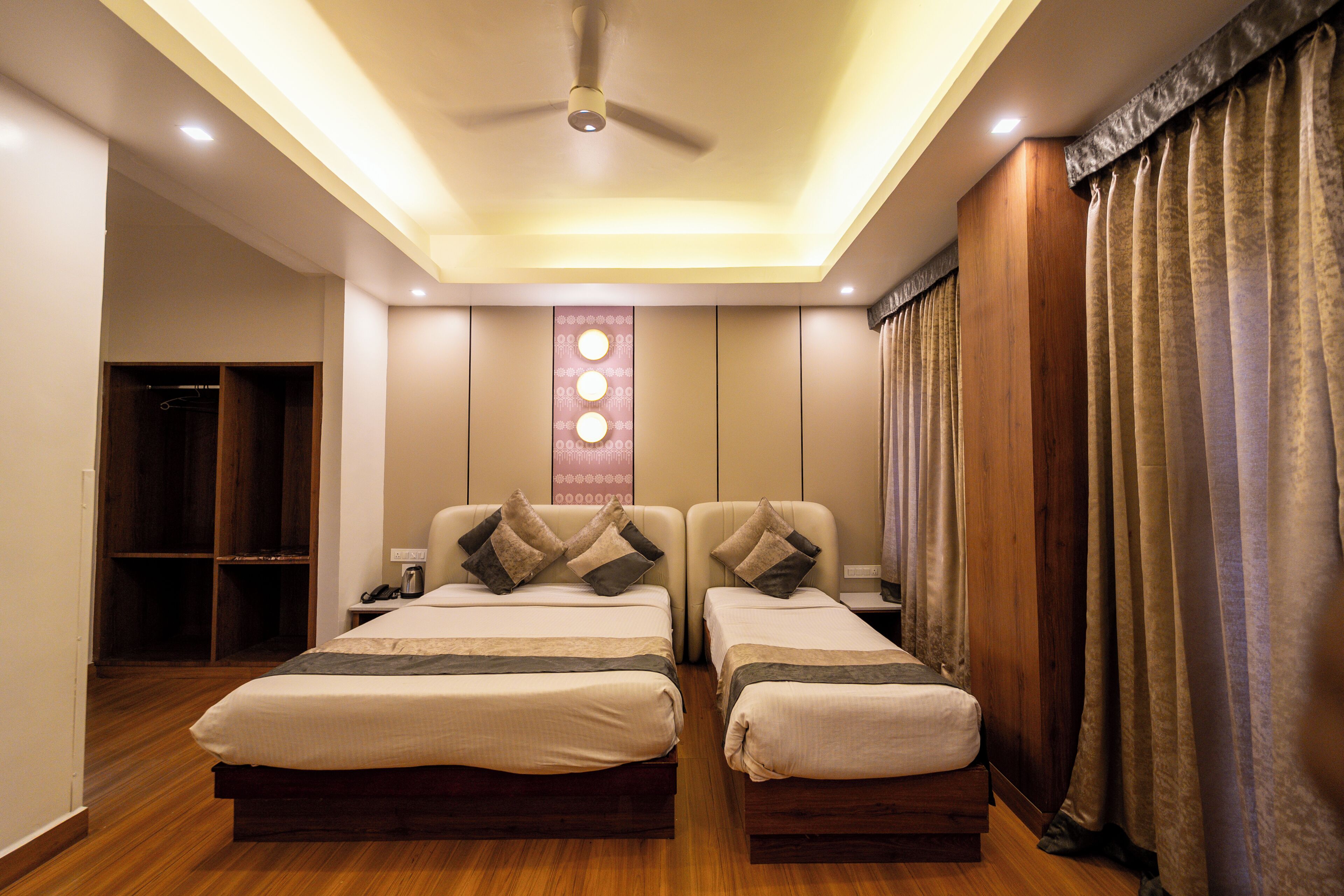 Quarto de hotel com duas camas, um ventilador de teto, um guarda-roupa de madeira e uma luminária na parede.