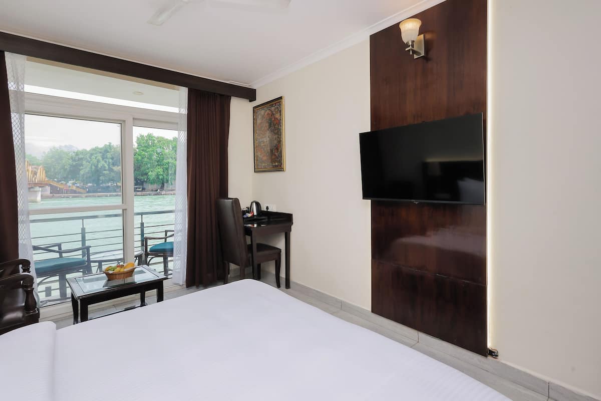 Quarto de hotel com cama, escrivaninha e cadeira, televisão na parede e janela com vista para a água e vegetação.